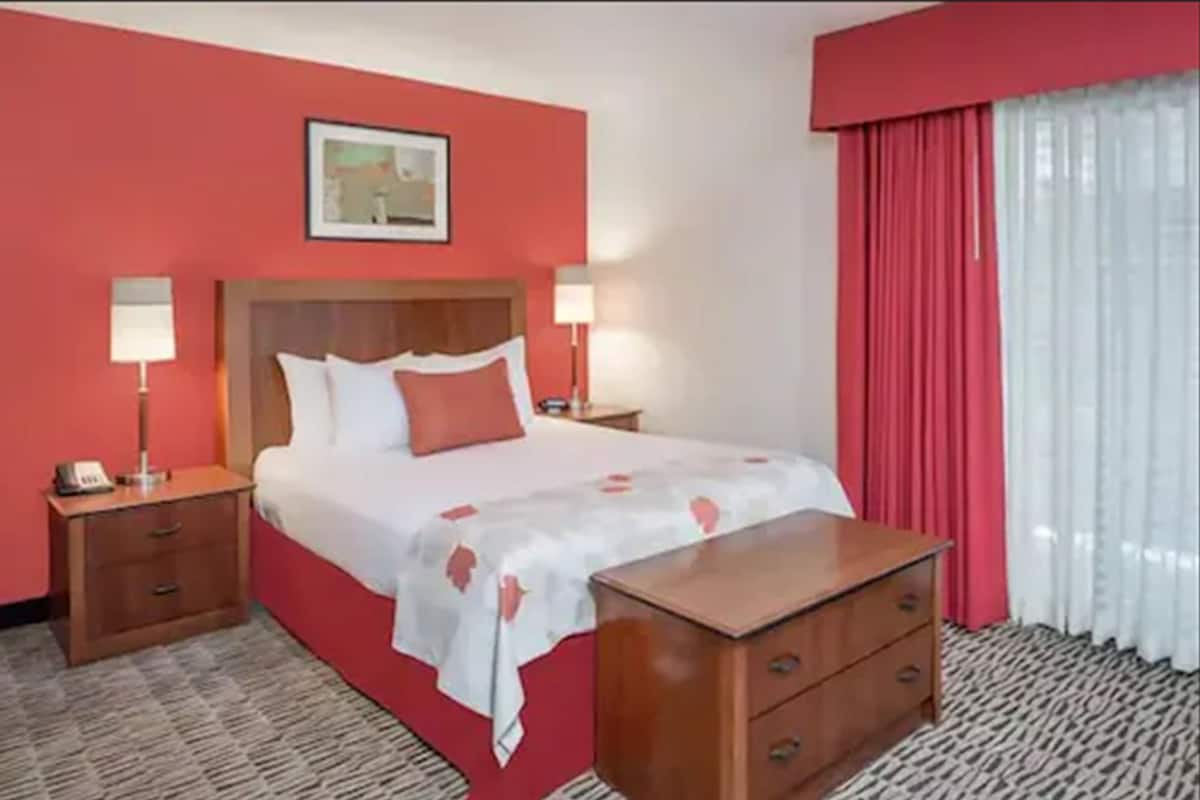Quarto de hotel com parede vermelha, cama de madeira com roupa de cama branca e vermelha, mesa de cabeceira com abajur e pequena cômoda de madeira.