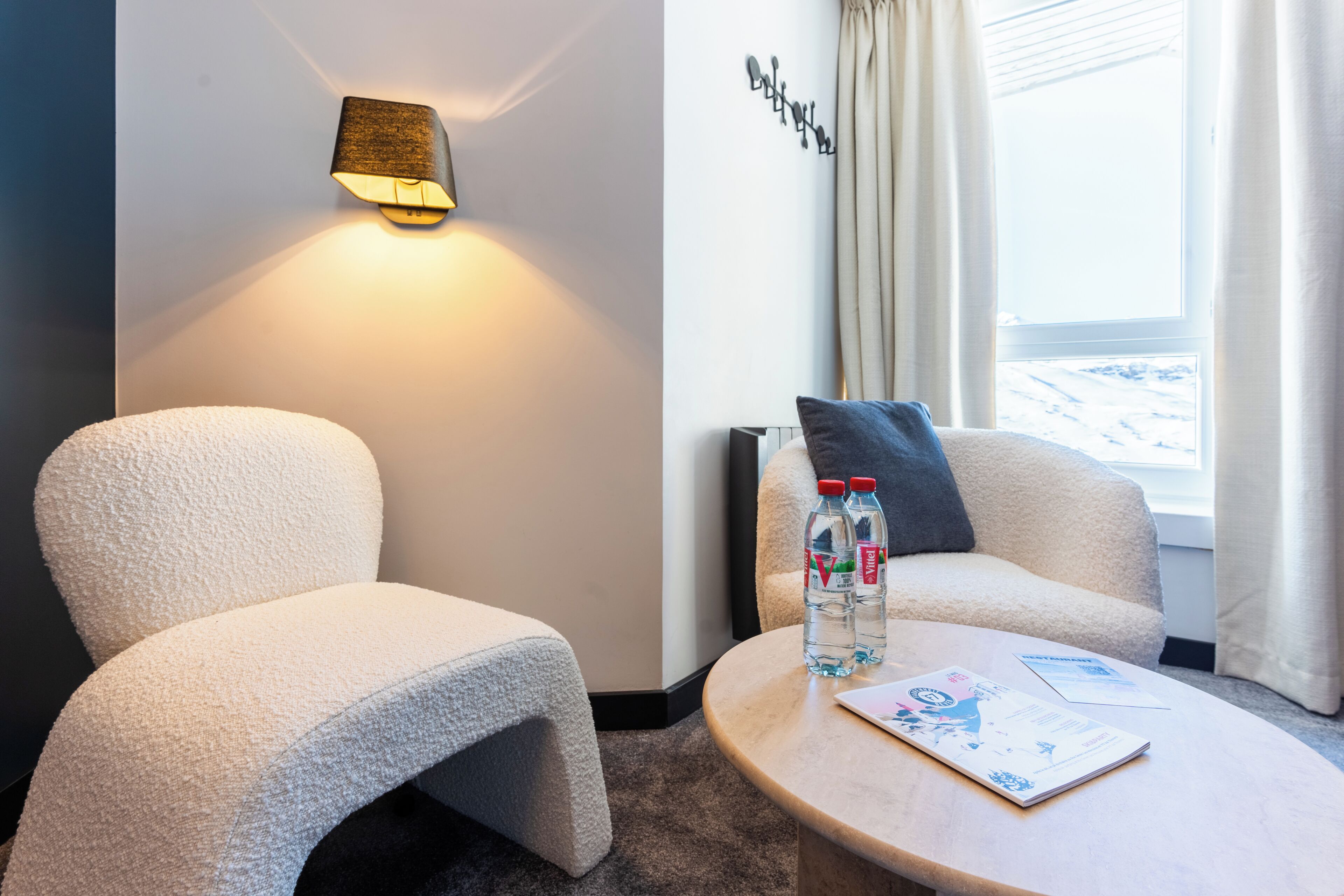 Quarto de hotel com duas poltronas confortáveis, uma mesa redonda com uma revista, uma garrafa de água e um abajur na parede.