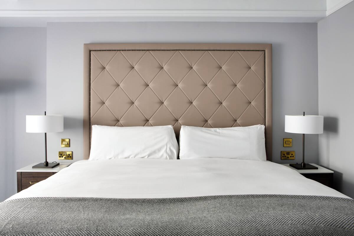 Cama bem arrumada com cabeceira estofada, duas mesas de cabeceira com abajures e um console lateral simples.