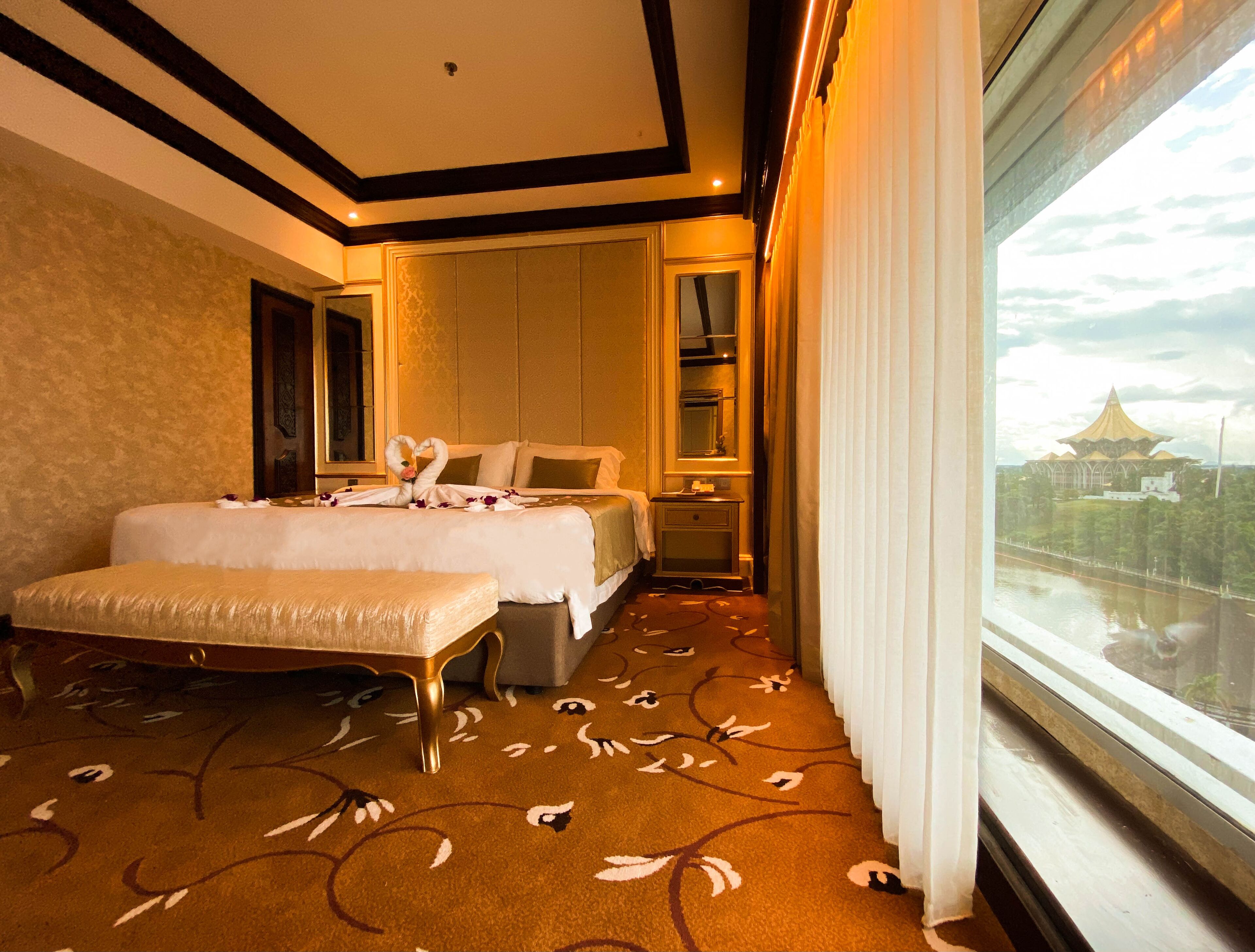 Quarto de hotel com uma cama grande, um espaço de estar, uma mesa de cabeceira e vista para um edifício distante pela janela.