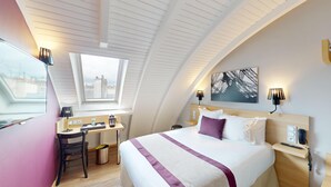 Quarto de hotel moderno com teto inclinado, uma mesa com cadeira, cama com cabeceira e janela com vista para a cidade.