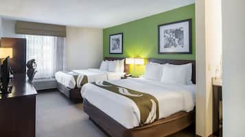 Quarto de hotel com duas camas, uma escrivaninha e uma cadeira. Há duas quadros nas paredes, um abajur na escrivaninha, e uma janela com cortinas.