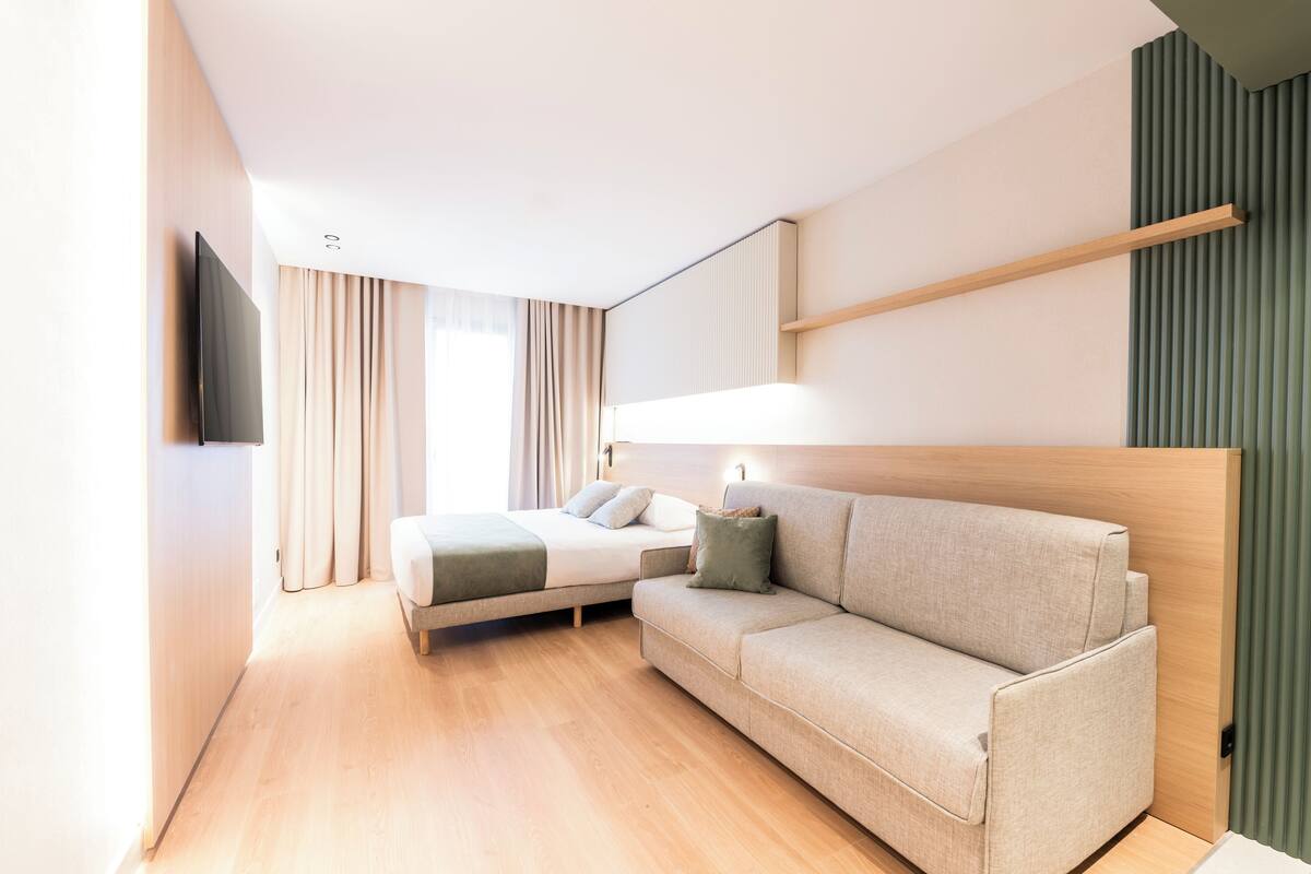 Quarto de hotel moderno com piso de madeira, sofá, cama, TV de tela plana e janela com cortinas.