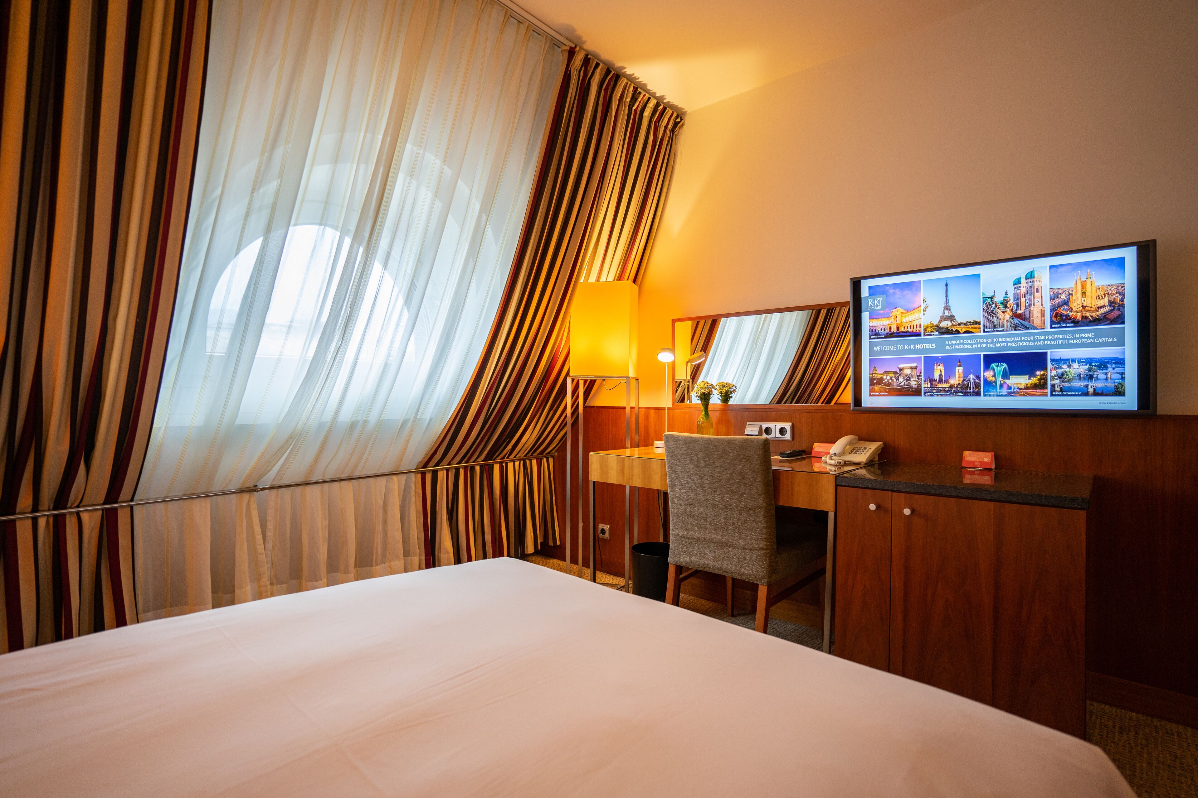 Quarto de hotel com uma cama grande, uma escrivaninha com cadeira, telefone e televisão na parede exibindo imagens de pontos turísticos famosos.