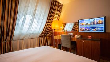 Quarto de hotel com uma cama grande, uma escrivaninha com cadeira, telefone e televisão na parede exibindo imagens de pontos turísticos famosos.