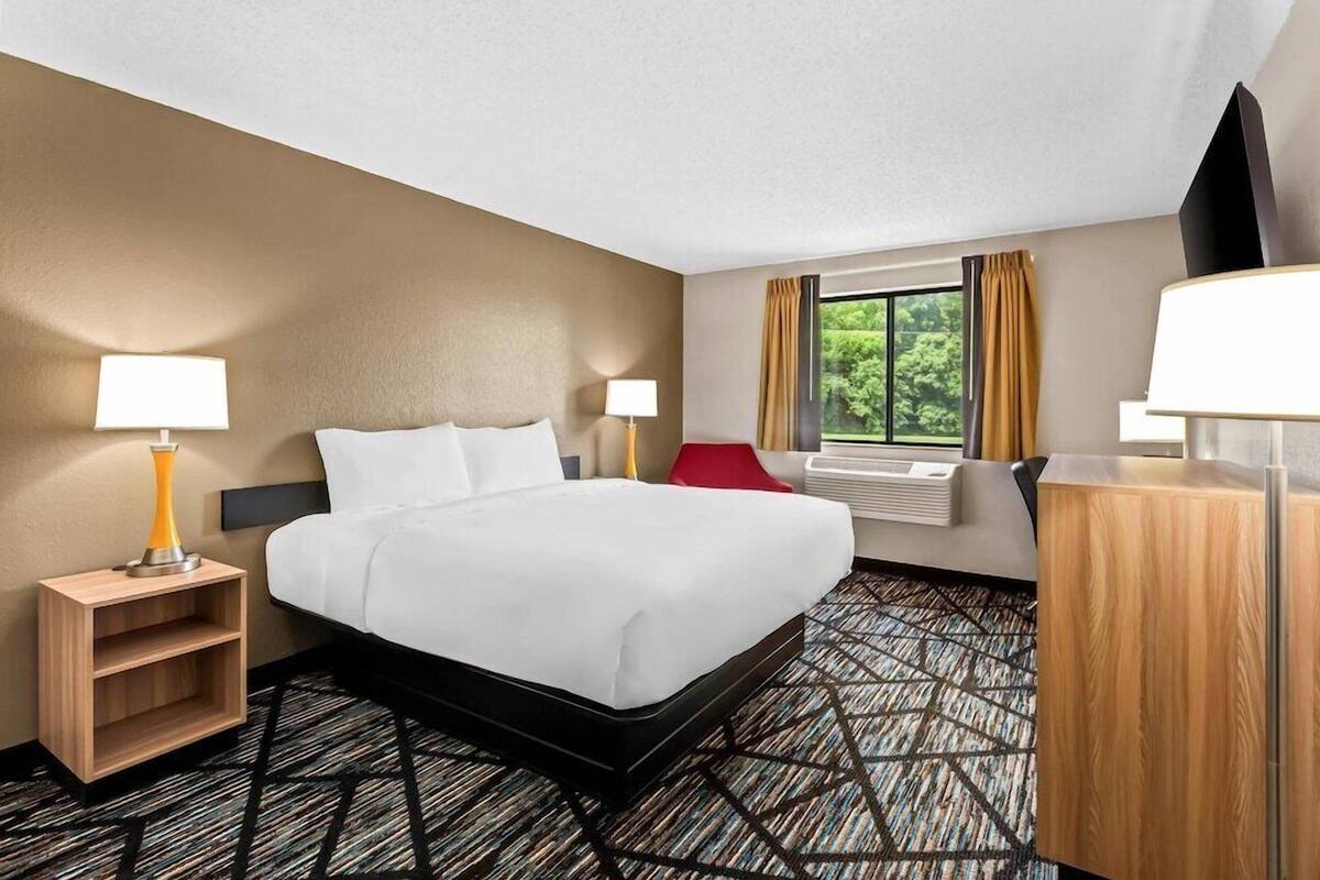 Quarto de hotel com cama, luminárias de cabeceira, escrivaninha e janela com cortinas.