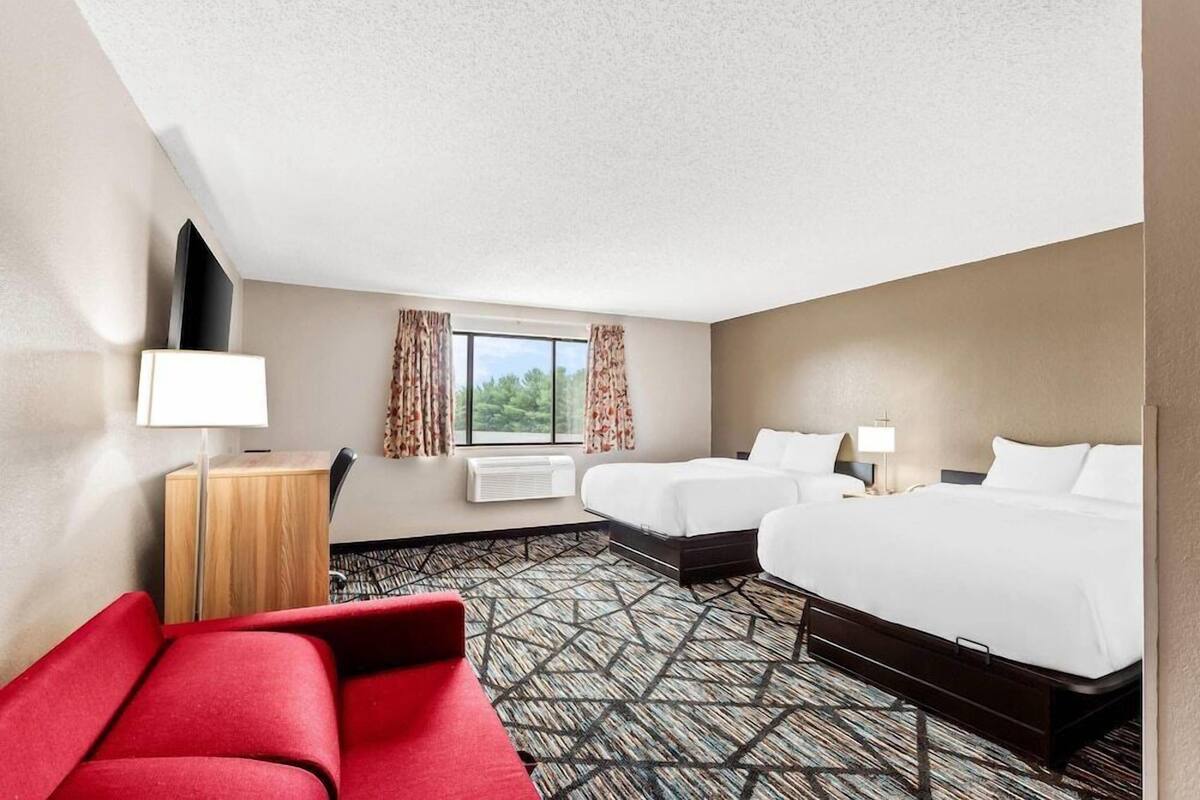 Quarto de hotel com duas camas, um sofá vermelho, uma televisão e uma janela com cortinas.