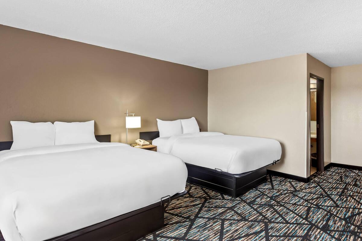 Quarto de hotel com duas camas, uma mesa de cabeceira com abajur e um tapete estampado.