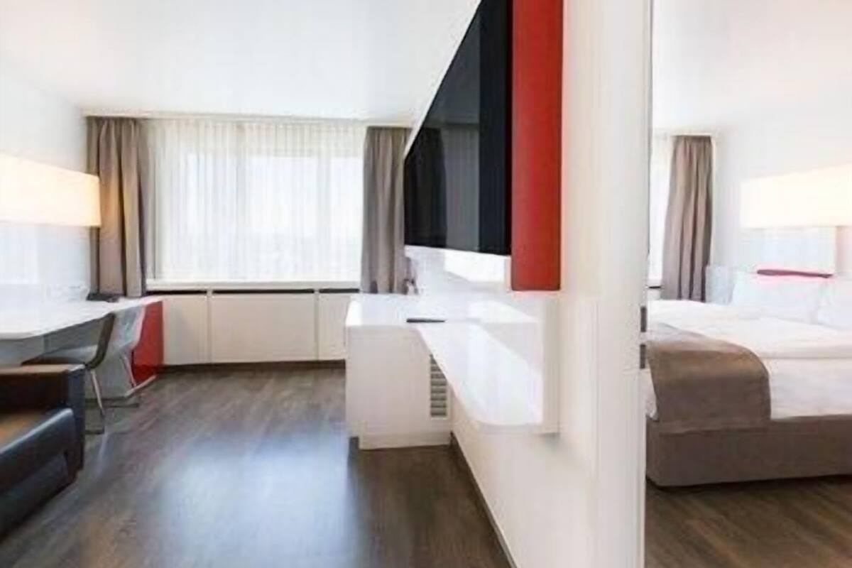 Quarto de hotel com cama, escrivaninha, cadeira, janela com cortinas e uma parede de destaque vermelha.