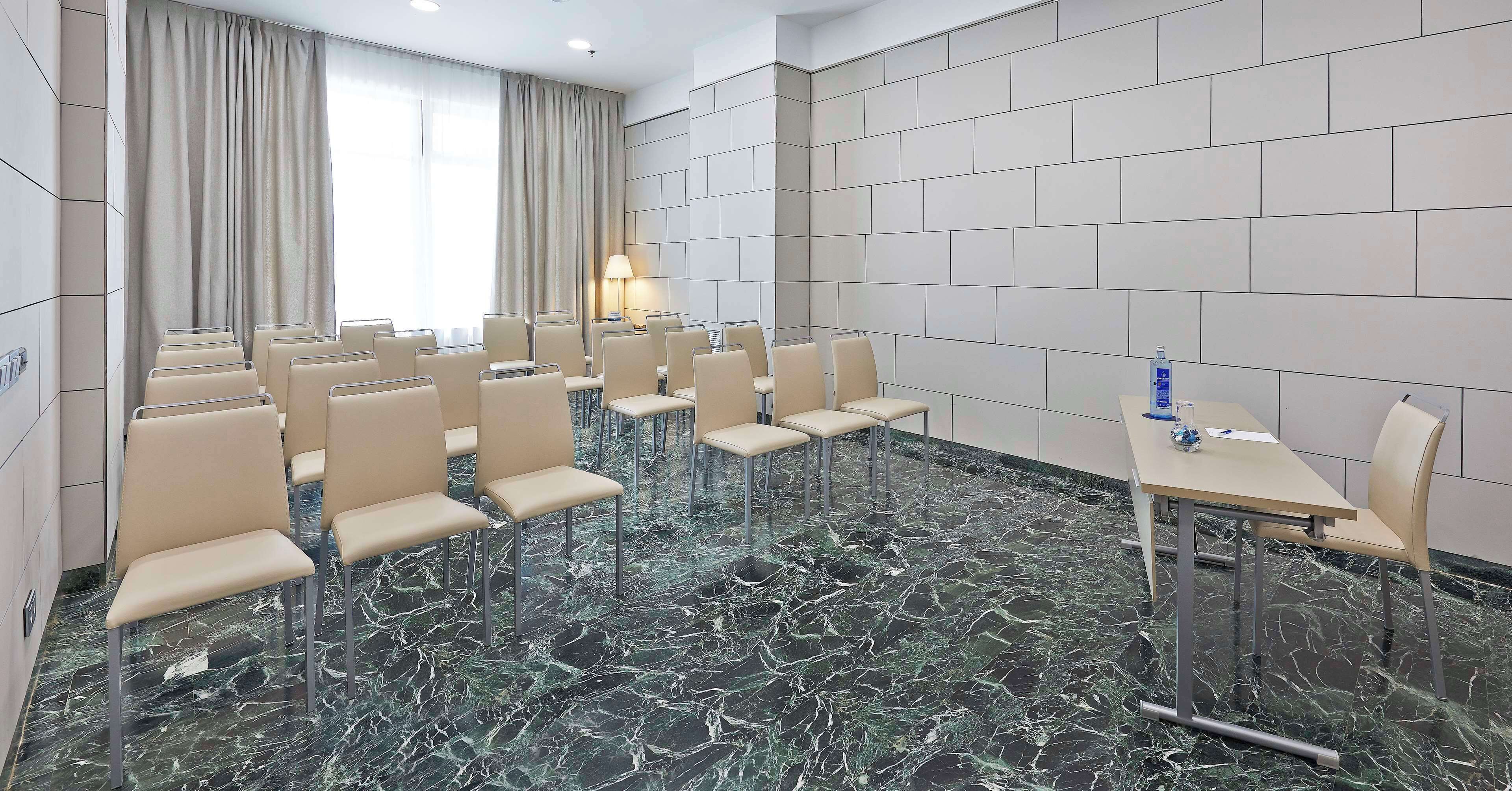Uma sala de conferências com piso de mármore, cadeiras bege, uma mesa com abajur e uma janela com cortinas.