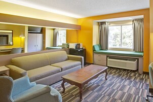 Quarto de hotel com sofá, mesa pequena, micro-ondas, escrivaninha, cadeira, janela com cortinas e espelho.