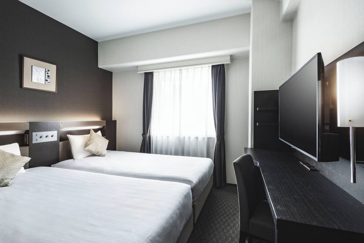 Quarto de hotel com duas camas, uma escrivaninha, televisão de tela plana e janela com cortinas.