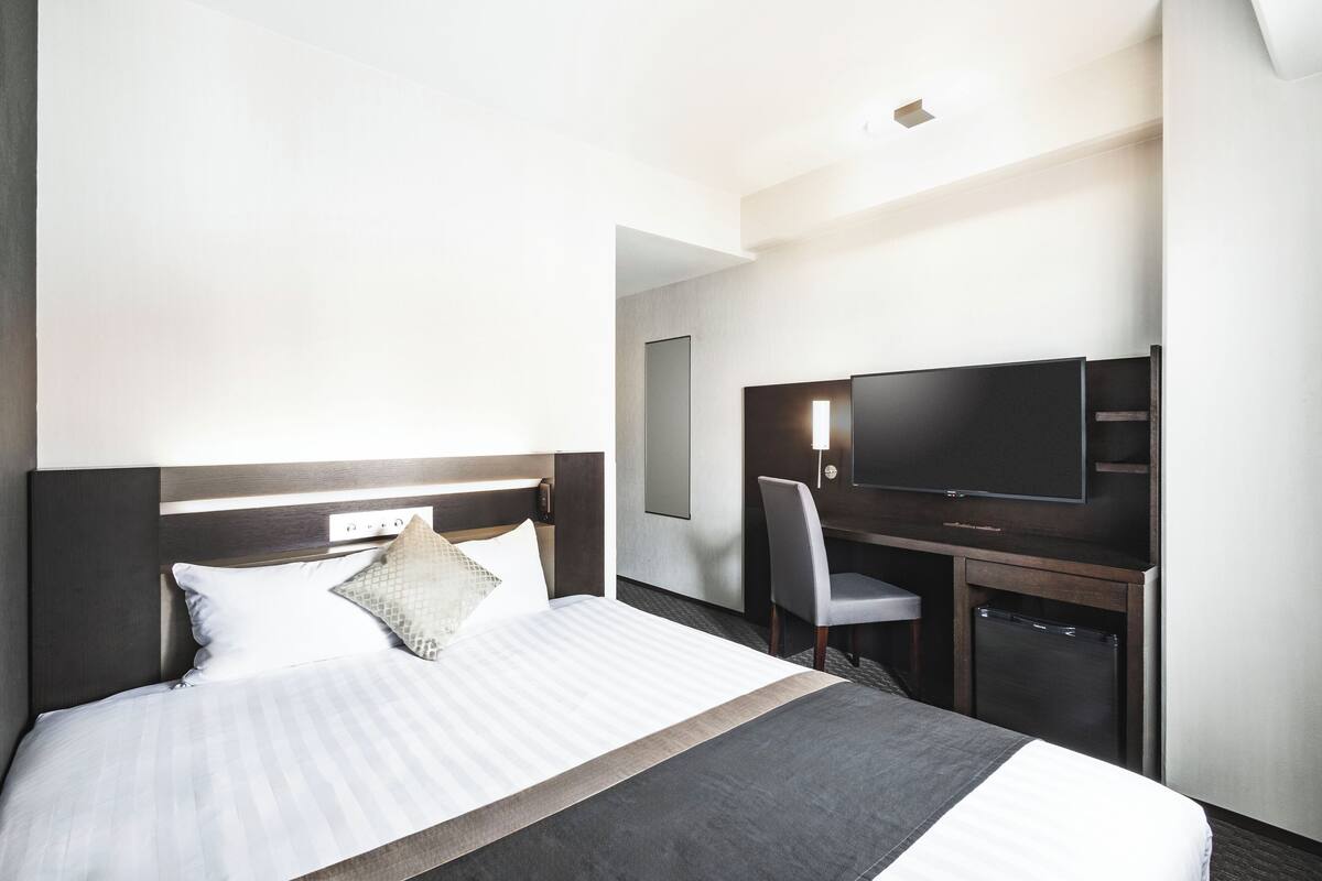 Quarto de hotel com uma cama grande, uma escrivaninha e uma televisão de tela plana, além de uma cadeira.