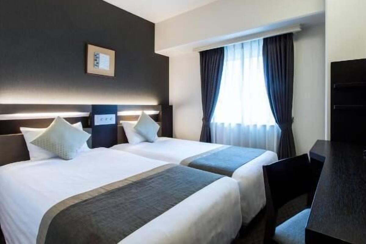 Quarto de hotel com duas camas, uma escrivaninha e uma janela com cortinas.