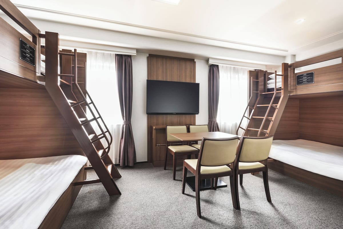 Quarto de hotel com beliches, televisão de tela plana, mesa de jantar com cadeiras e piso coberto por carpete.