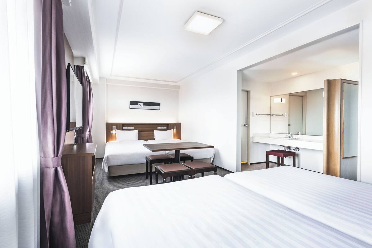 Quarto de hotel com duas camas, uma escrivaninha, uma cadeira, uma mesinha, um espelho e banheiro com pia e espelho.