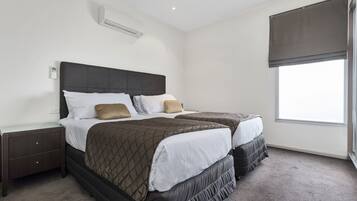 Quarto com duas camas, mesa de cabeceira, ar condicionado fixado na parede e uma janela grande com cortina.