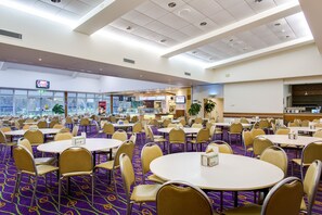 Sala de conferências com mesas e cadeiras redondas, área de buffet e tela de televisão.