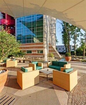 Uma área de estar ao ar livre com móveis modernos, um grande vaso de plantas e um edifício com janelas de vidro refletivo ao fundo.