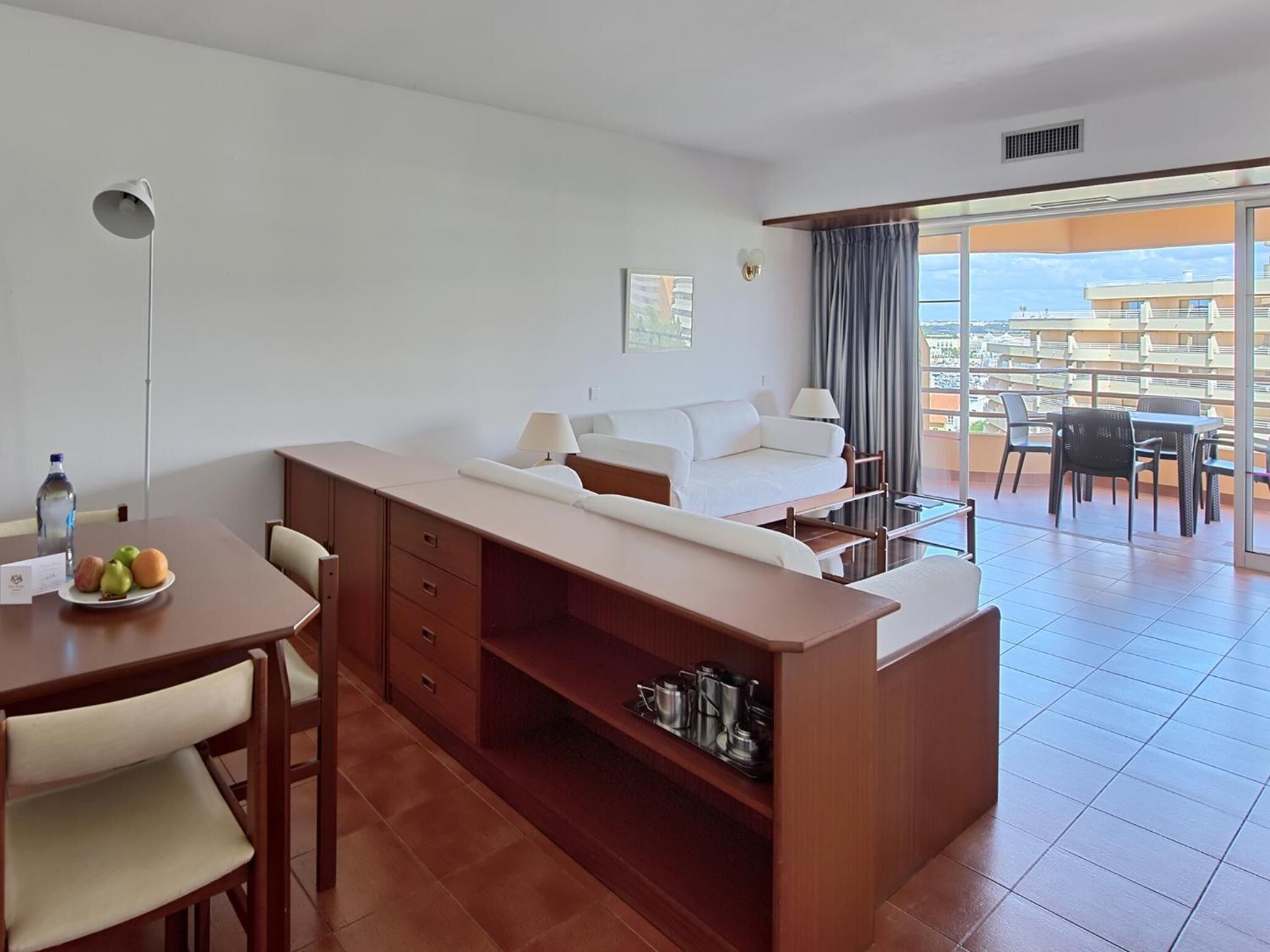 Quarto de hotel com área de refeições, sofá-cama, varanda com mesa e cadeiras, e vista para a cidade.