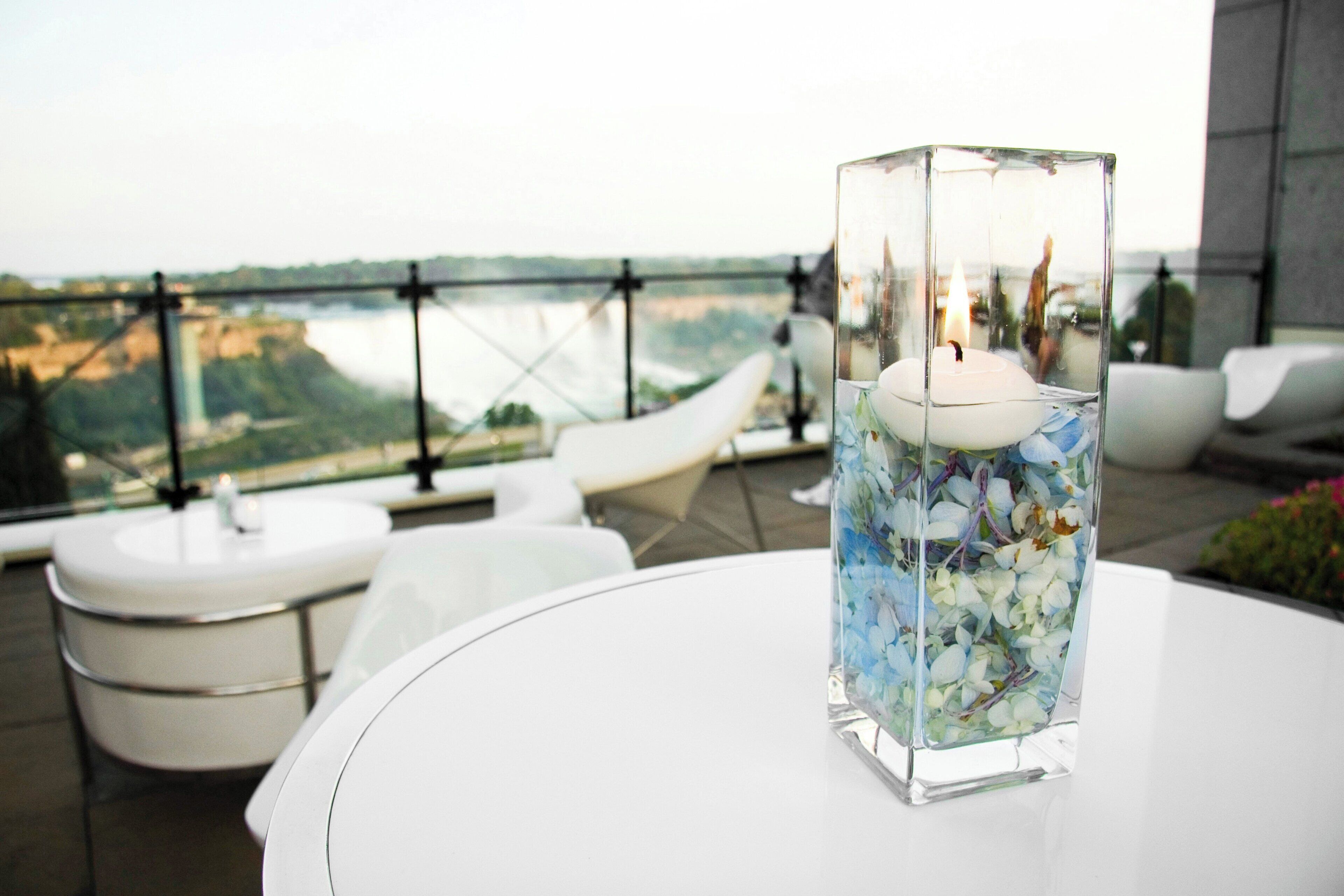 Um vaso de vidro transparente com flores sobre uma mesa branca numa varanda com vista panorâmica.