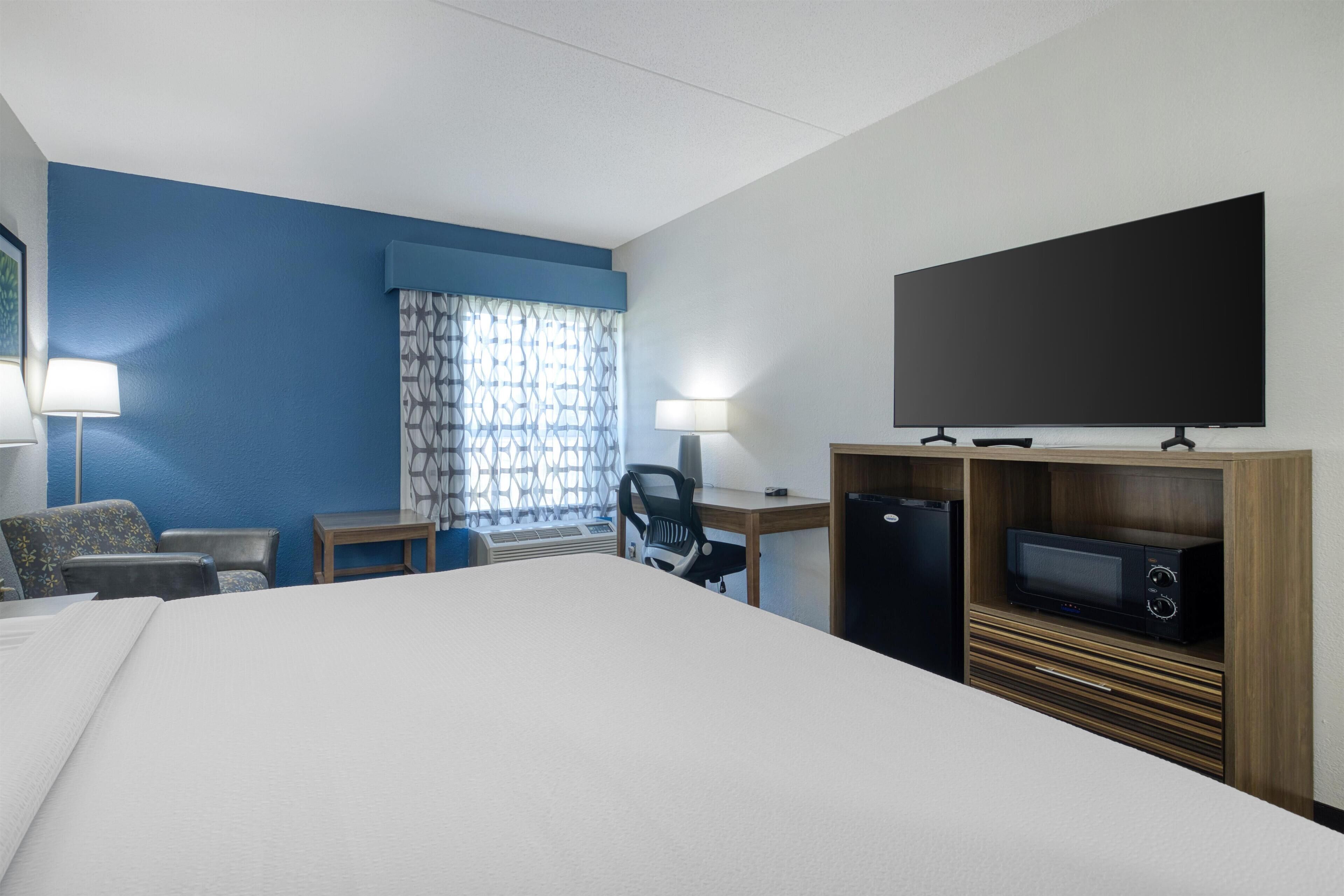 Quarto de hotel com uma cama grande, uma escrivaninha com computador, televisão, micro-ondas e uma cadeira.
