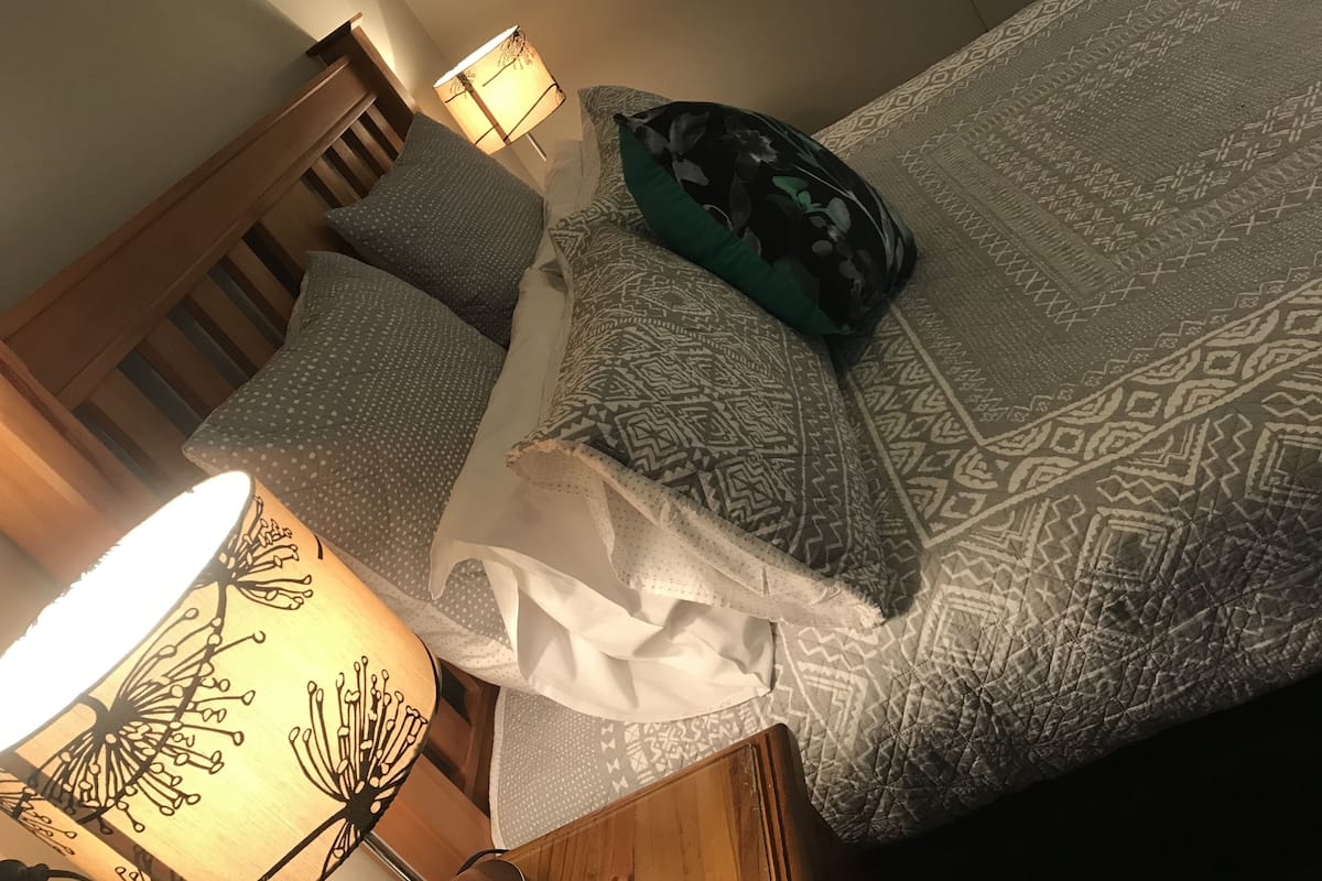 Cama bem arrumada com roupa de cama estampada e almofadas, abajur e cabeceira de madeira.