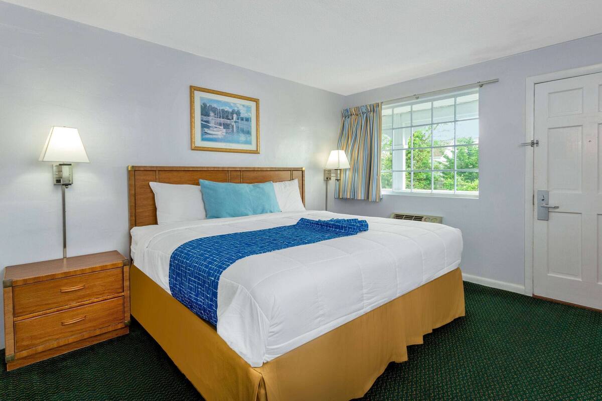 Cama bem arrumada com colcha azul e branca, mesa de cabeceira de madeira com gavetas, quadro na parede e janela com cortinas.