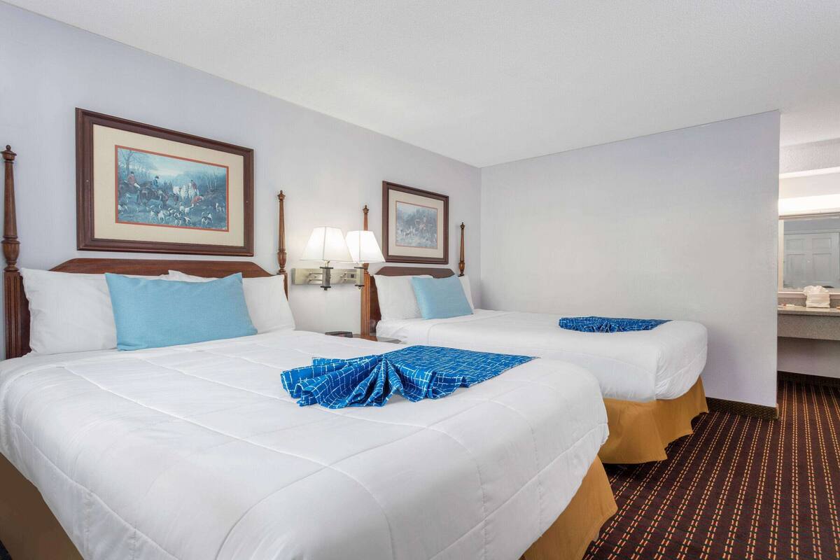 Quarto de hotel com duas camas, roupa de cama branca, almofadas azuis e um objeto decorativo azul sobre as camas. Há quadros nas paredes, uma mesa de cabeceira com uma lâmpada e um banheiro visível no canto.