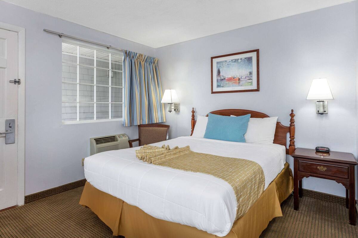 Quarto de hotel com uma cama grande, duas mesas de cabeceira, uma janela com cortinas, um quadro na parede e uma porta.