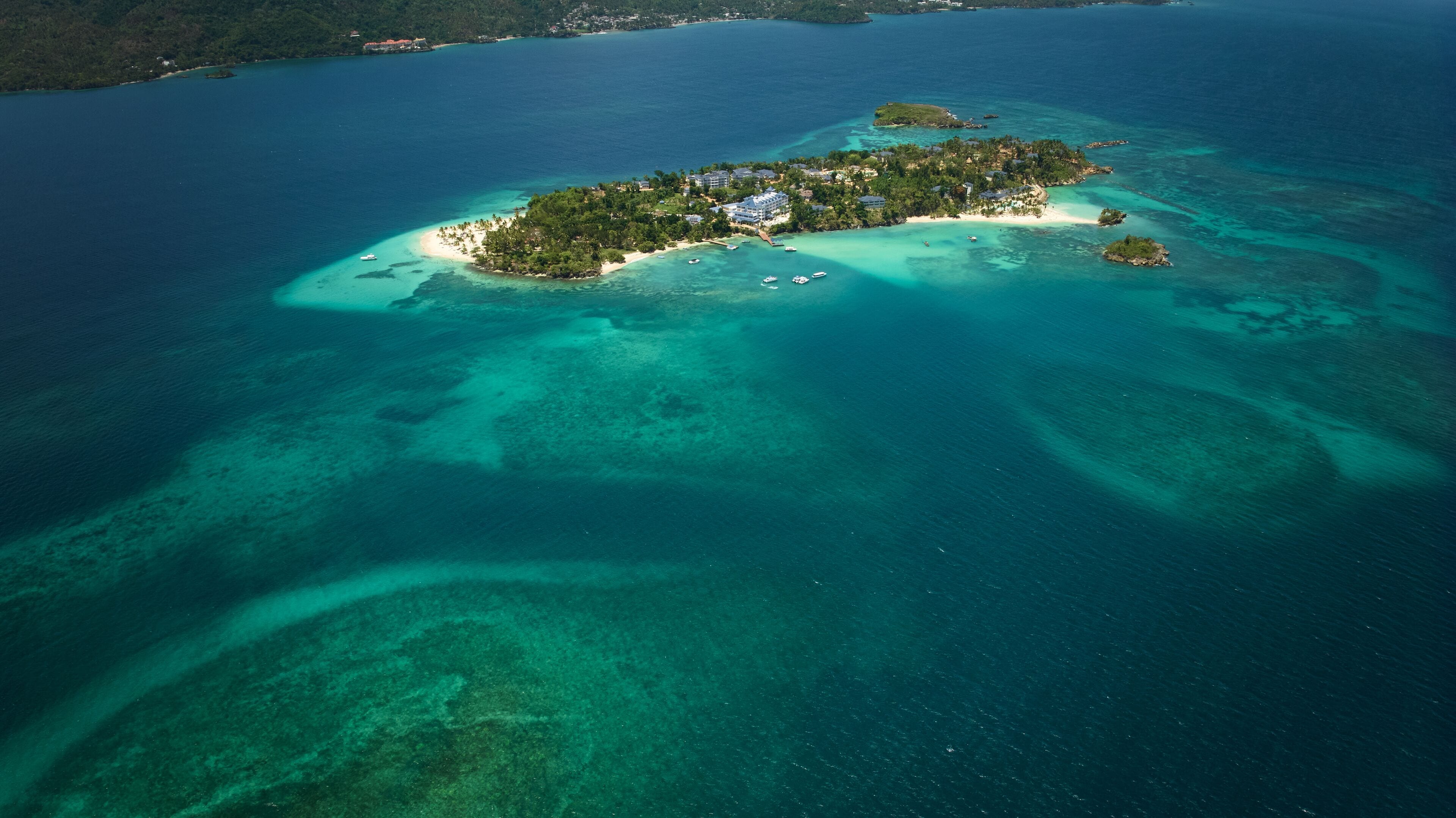 Vista aérea de uma ilha tropical com águas turquesa cristalinas, cercada por vegetação exuberante e alguns edifícios.