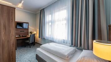Quarto de hotel com uma cama, uma escrivaninha com cadeira, televisão e janela com cortinas.
