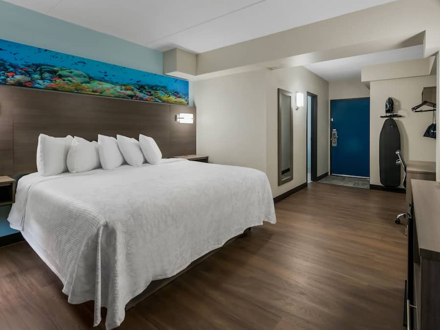 Quarto de hotel com cama, mesa de cabeceira, relógio de parede, pintura de uma cena subaquática e porta para outro cômodo.