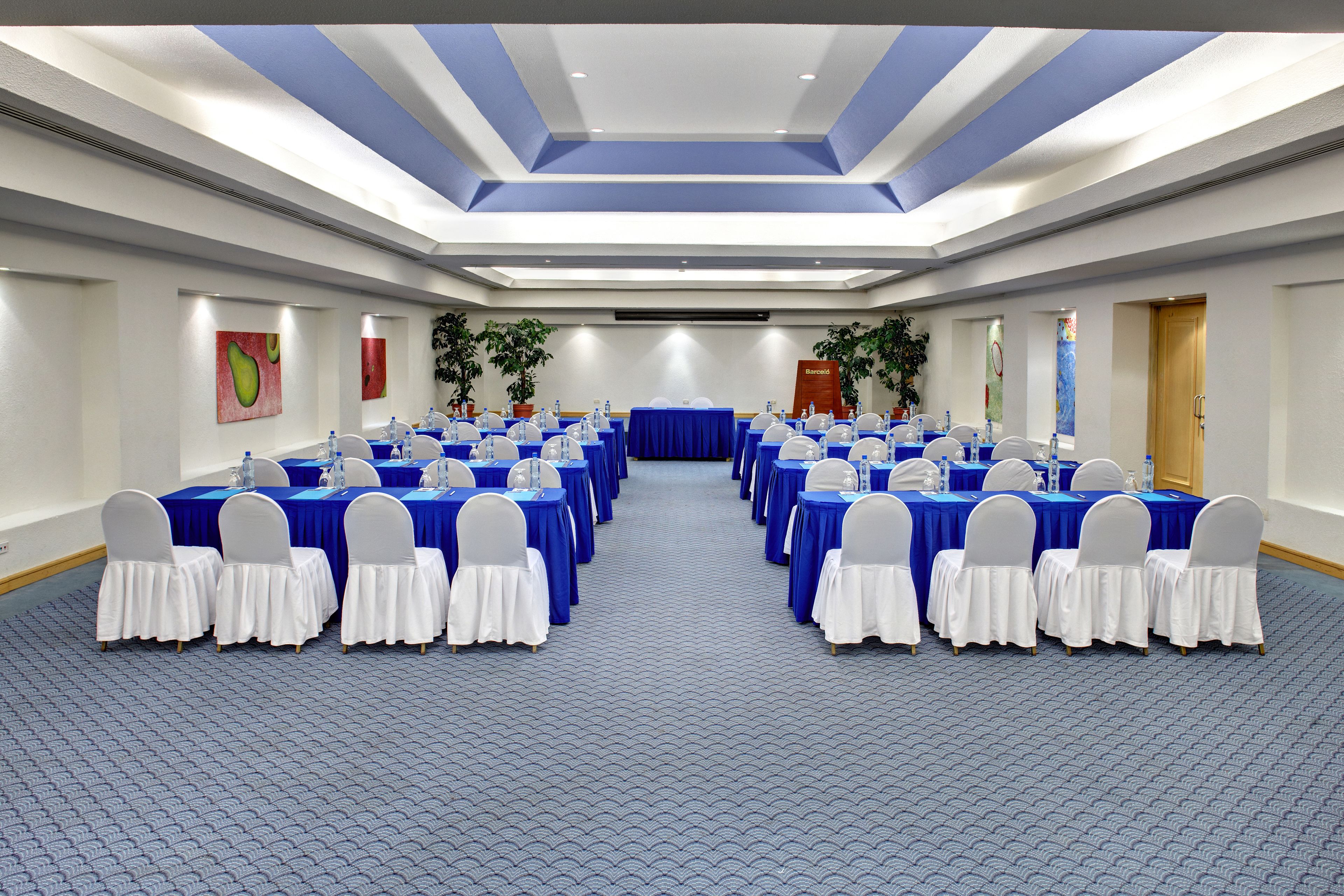 Uma sala de conferências com fileiras de mesas e cadeiras, carpete azul e design de teto moderno.