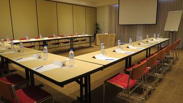 Sala de conferência com mesas compridas, cadeiras, garrafas de água, placas de identificação e uma tela de projetor.