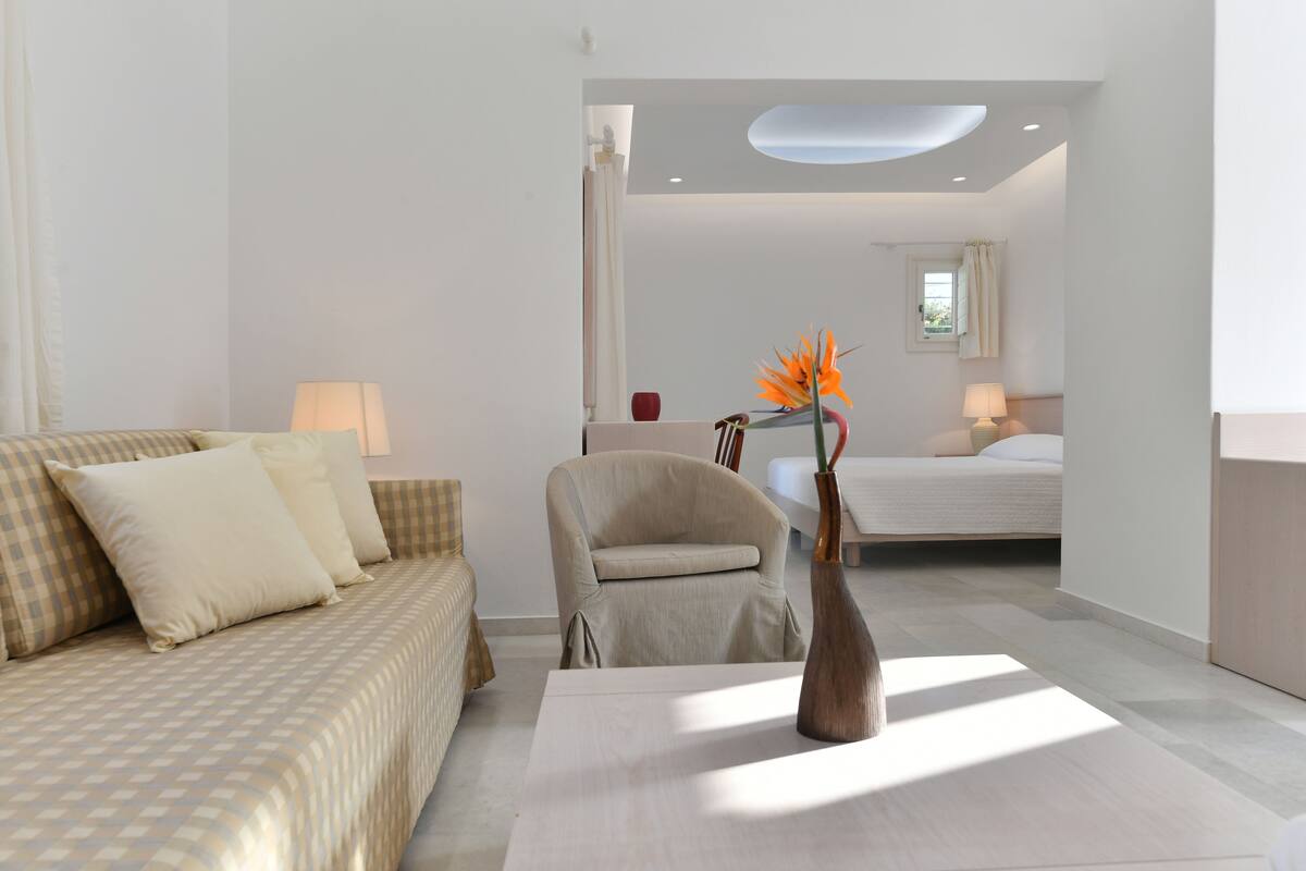 Quarto de hotel moderno com sofá, poltrona, mesa de jantar e um vaso com flores.