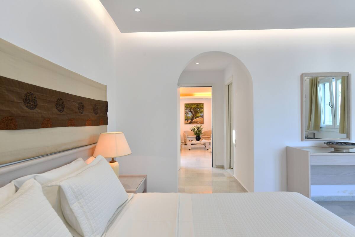 Quarto de hotel com cama, mesa de cabeceira, luminária, pintura na parede, escrivaninha com espelho e porta em arco que dá para outro cômodo.