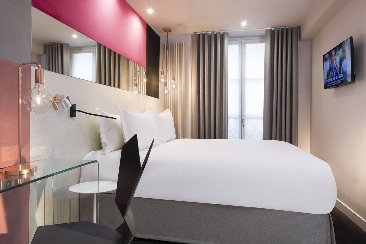 Quarto de hotel moderno com uma cama grande, uma mesa de vidro, uma escrivaninha e uma televisão na parede.