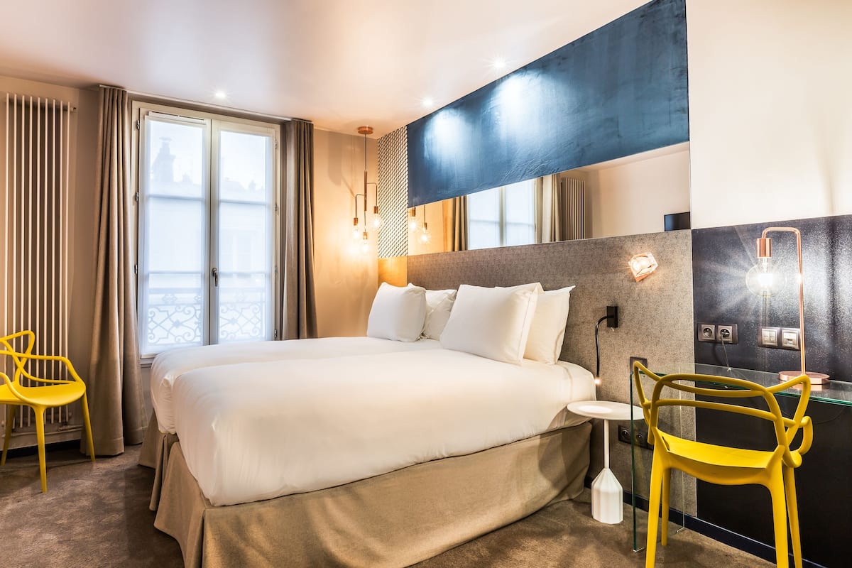 Quarto de hotel com uma cama grande, uma mesa de vidro, uma cadeira amarela e vista para a cidade pela janela.