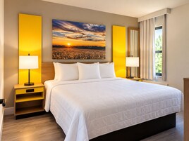 Quarto de hotel com uma cama grande, duas mesas de cabeceira com luminárias, um criado-mudo, uma janela ampla com cortinas e uma pintura de pôr do sol na parede.