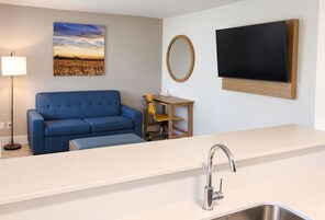 Uma cozinha compacta moderna com pia, um sofá azul, uma pequena mesa com uma cadeira, uma TV fixada na parede e uma pintura de um campo.