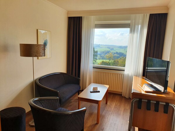 Quarto de hotel com sofá, uma mesa pequena, televisão e janela com vista para o campo.