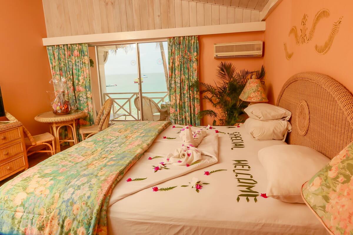 Quarto com uma cama, uma cadeira de vime, uma mesinha e vista para o mar através de uma porta de correr.
