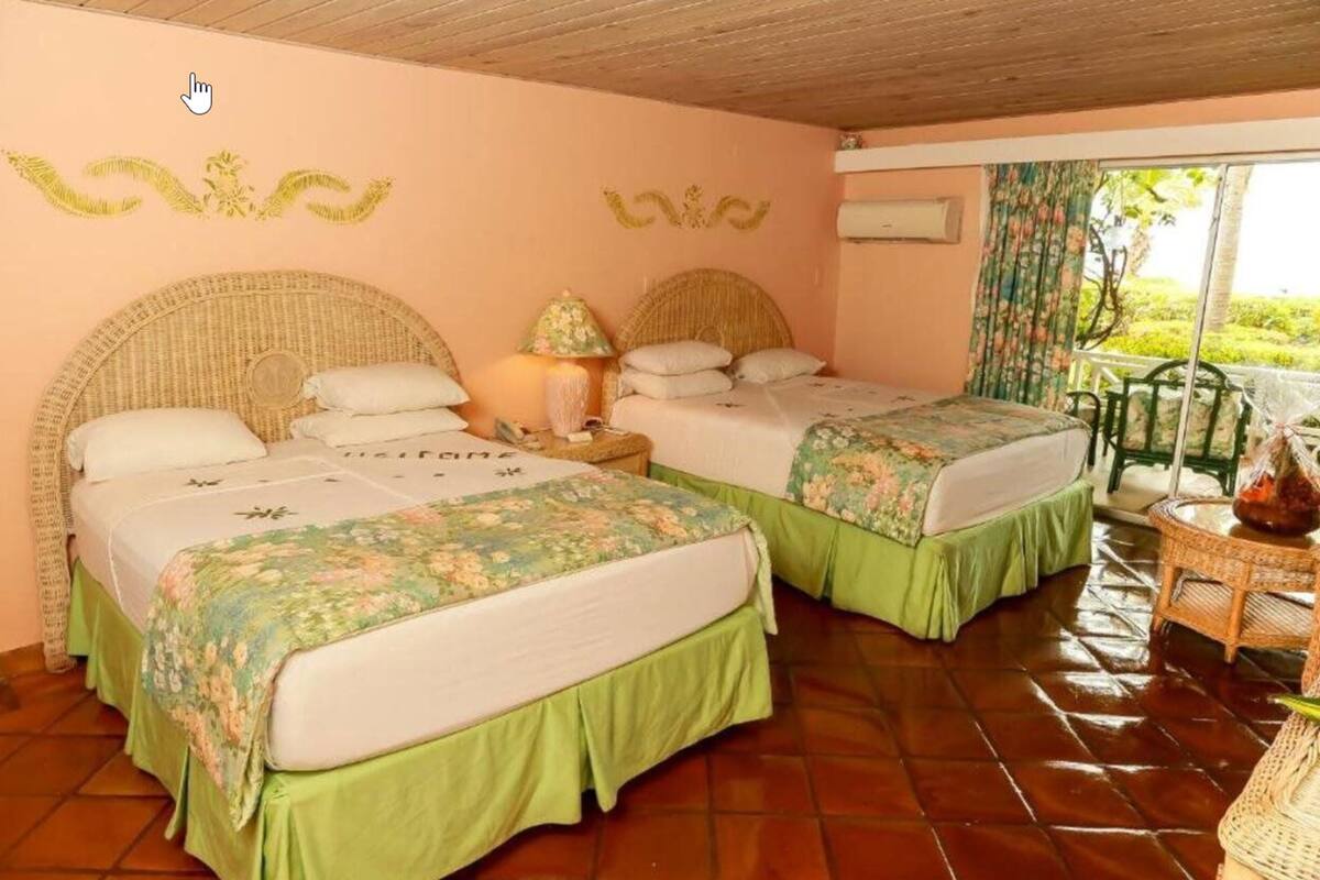 Quarto com duas camas, cada uma com saia verde e colcha floral. Há uma cadeira de vime e uma mesa pequena com abajur.