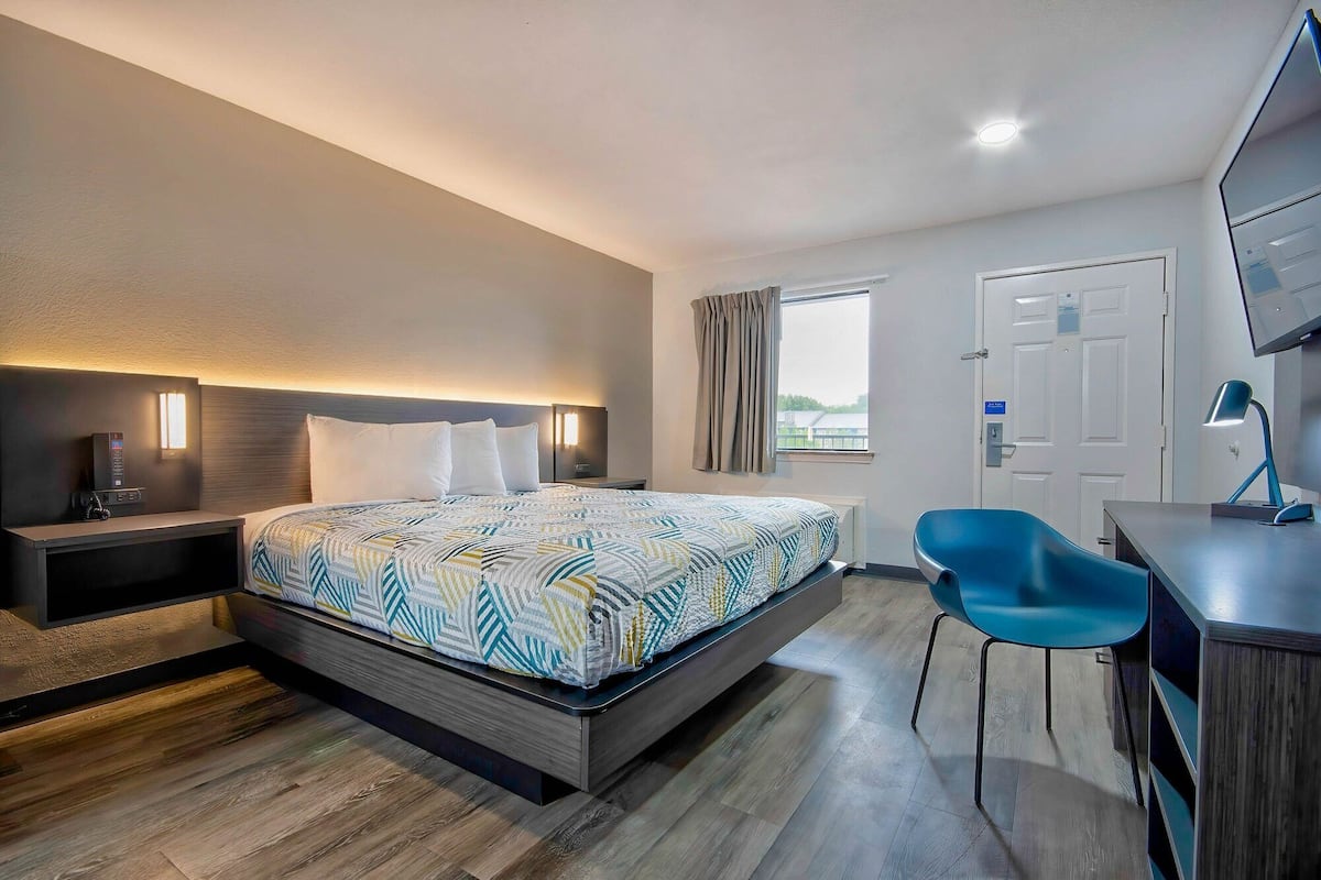 Quarto de hotel moderno com uma cama grande, mesas de cabeceira, escrivaninha com cadeira, televisão e janela com cortinas.