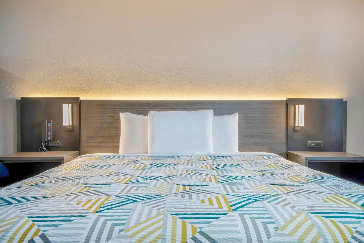 Quarto de hotel com uma cama, duas mesas de cabeceira e duas luminárias fixadas na parede.