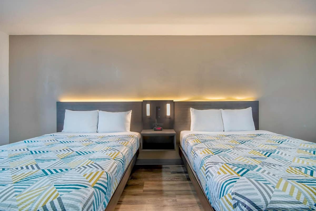 Quarto de hotel com duas camas de solteiro, cada uma com colcha estampada, mesa de cabeceira com relógio e luminária fixada na parede entre as camas.