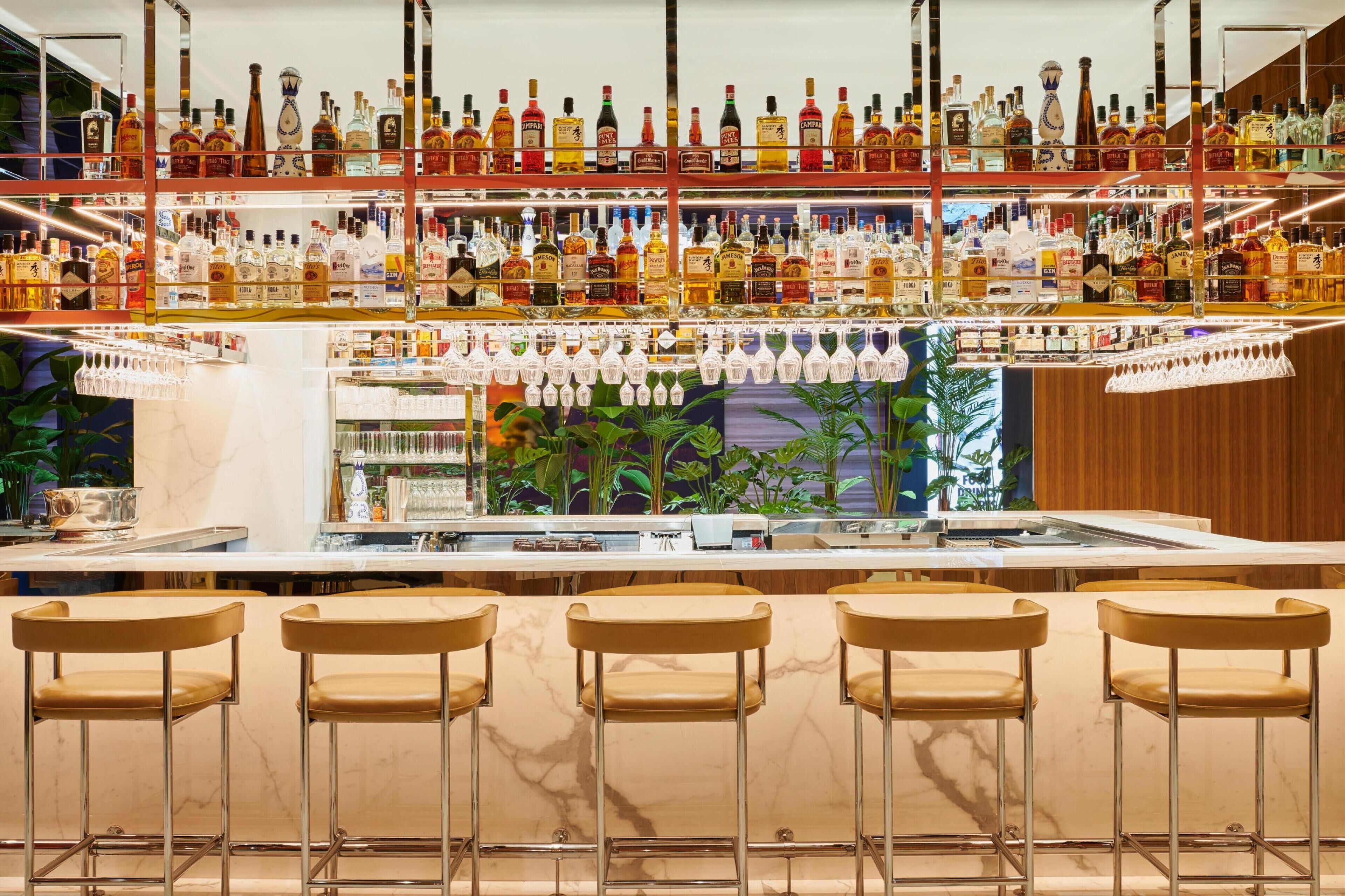 Uma área de bar com várias prateleiras de garrafas de bebidas, um balcão com banquetas e vista para uma parede de plantas verdes.