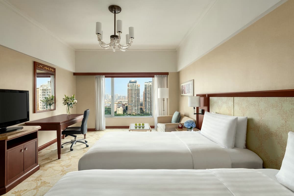 Quarto de hotel com uma cama grande, uma escrivaninha com televisão, uma cadeira, uma mesa pequena e vista da cidade por grandes janelas.