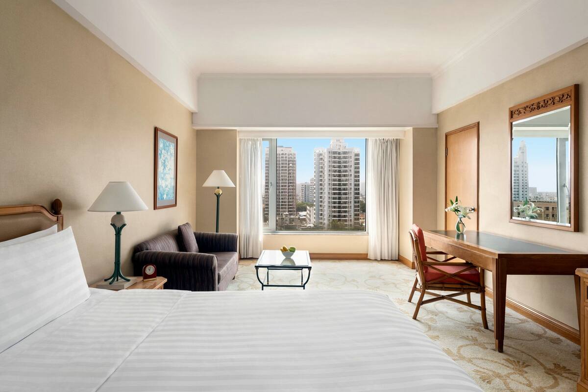 Quarto de hotel com uma cama grande, uma escrivaninha, uma cadeira, uma mesinha e vista para edifícios altos.